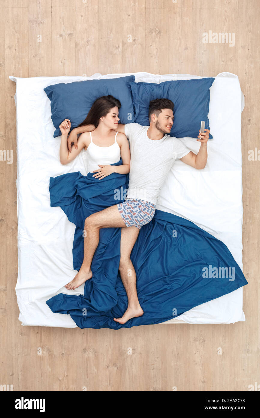 Bedtime. Young man browsing social media putting leg on sleeping woman ...