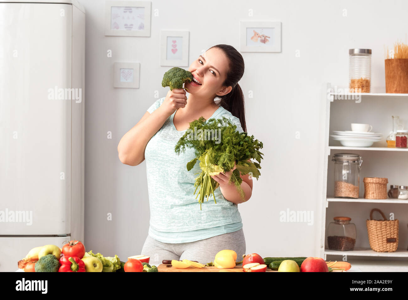 Cooking Meal. Chubby girl standing in kitchen with greenery biting ...