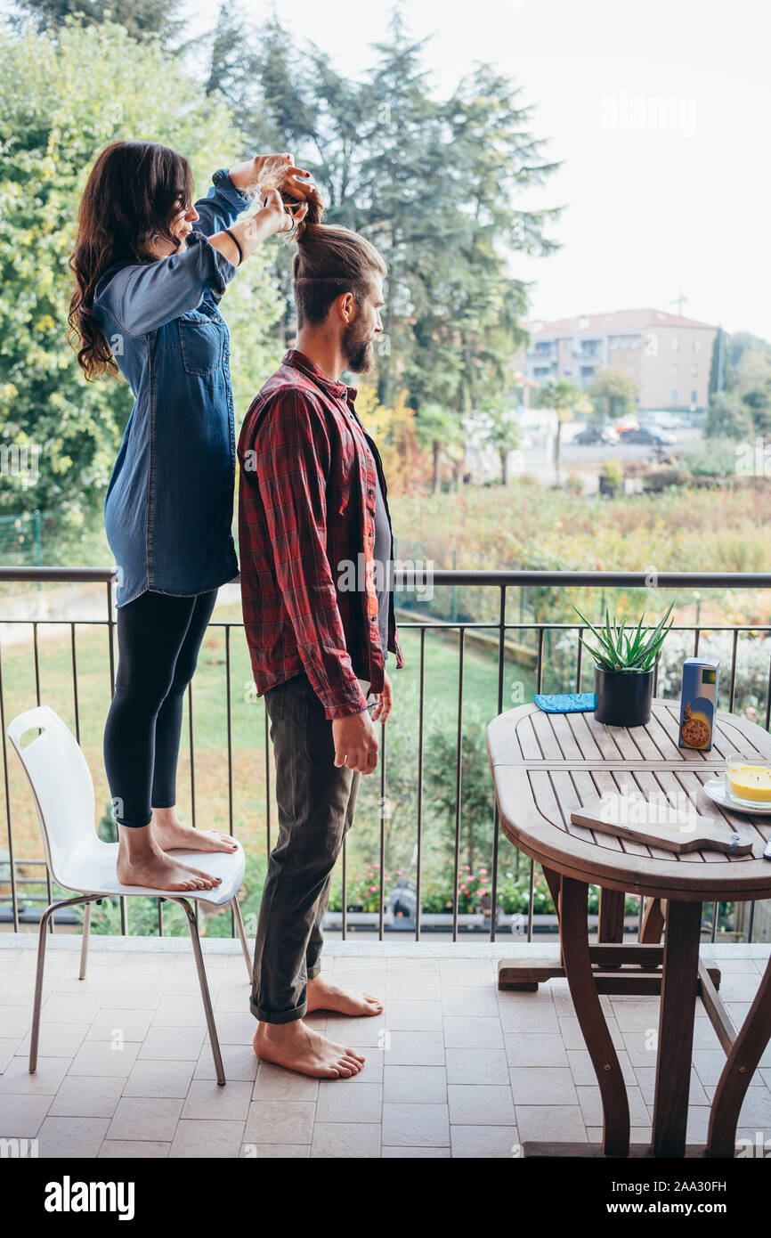 Young woman standing on a chair doing hair bun to her taller boyfriend ...