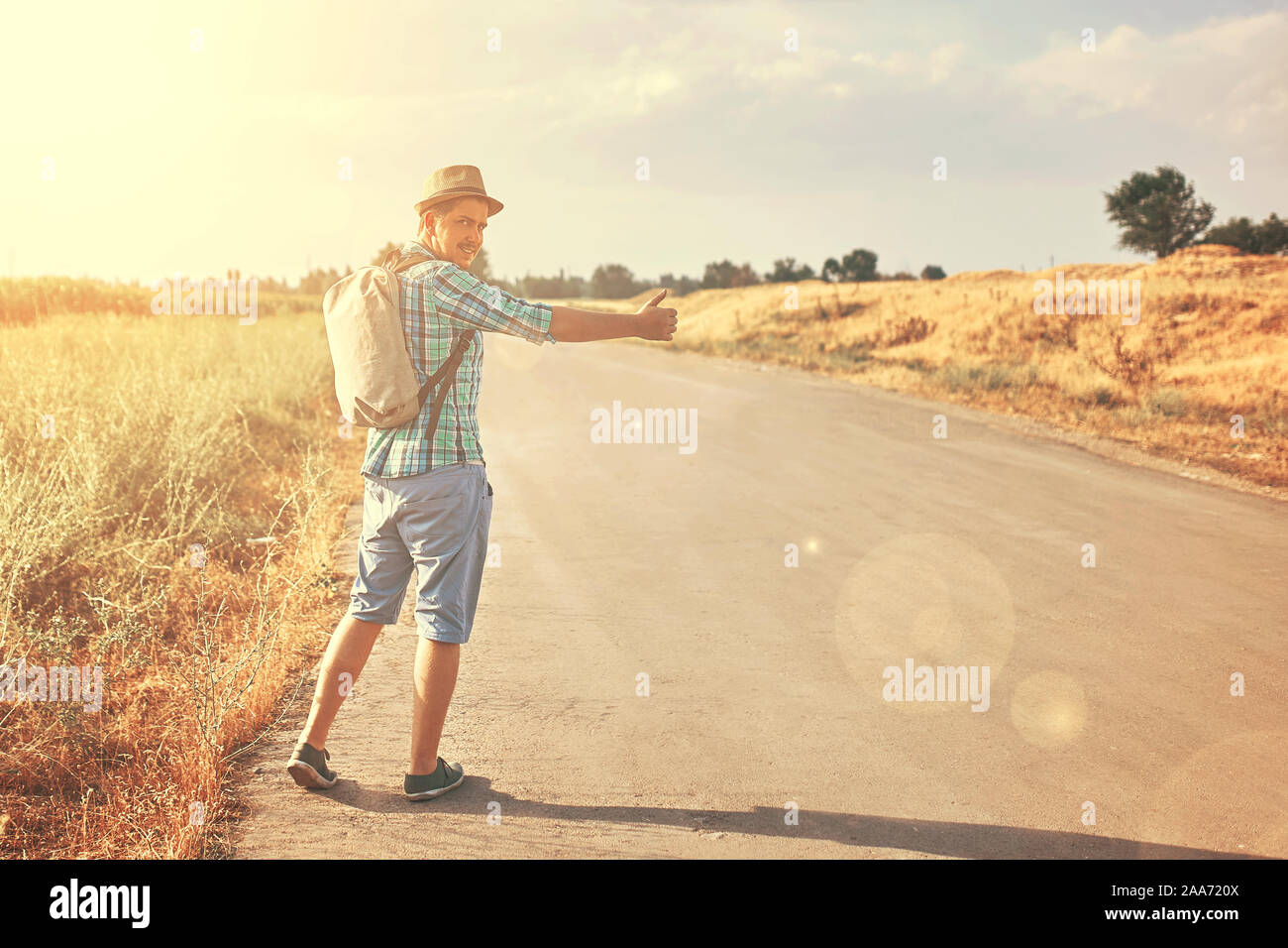 Hitchhiker with backpack in summer hat, light shirt, shorts on the road ...