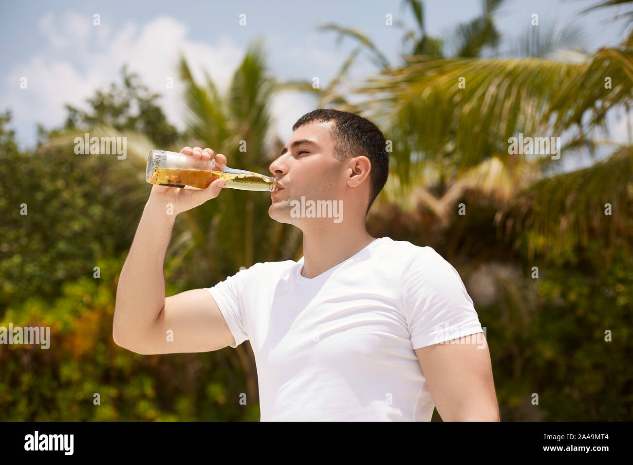 Young man drinking beer on the tropical beach with coconut palm trees ...