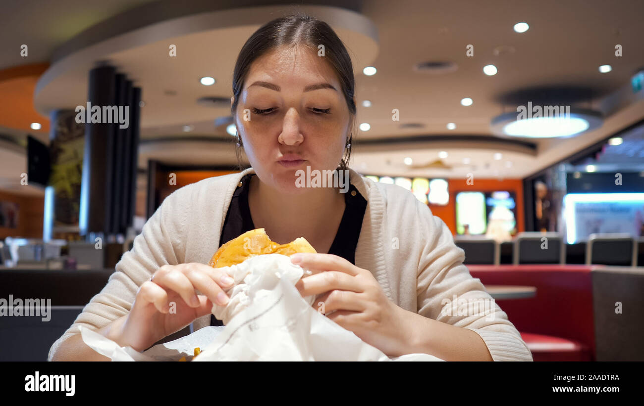 Junk food concept. Young brunette woman is eating burger in fast food ...