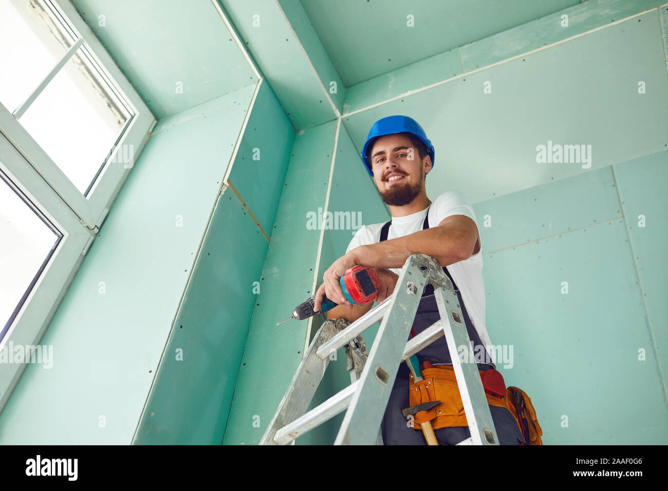 A builder standing on a ladder installs drywall at a construction site ...