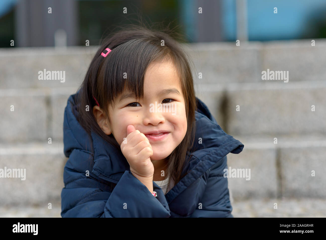 Cute smiling little Asian girl in a warm blue jacket giving a thumbs up ...