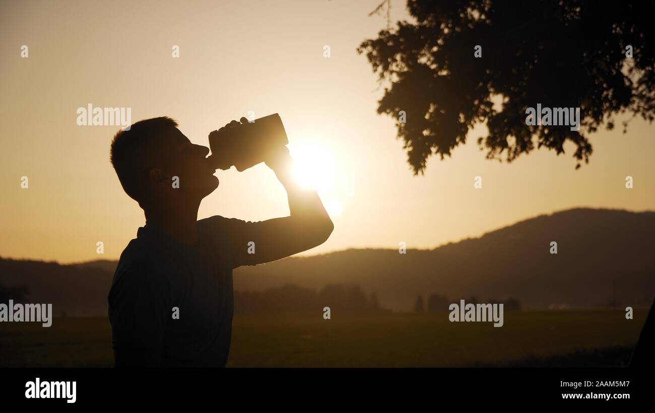 silhouette of fit young man drinking water with sun behind it in sunset ...