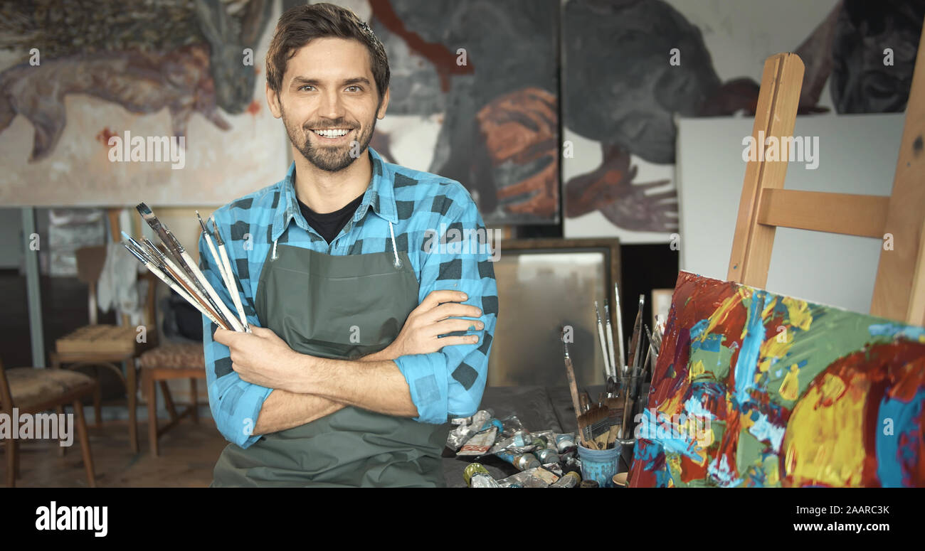 Portrait of young smiling artist posing with paintbrushes in his loft ...