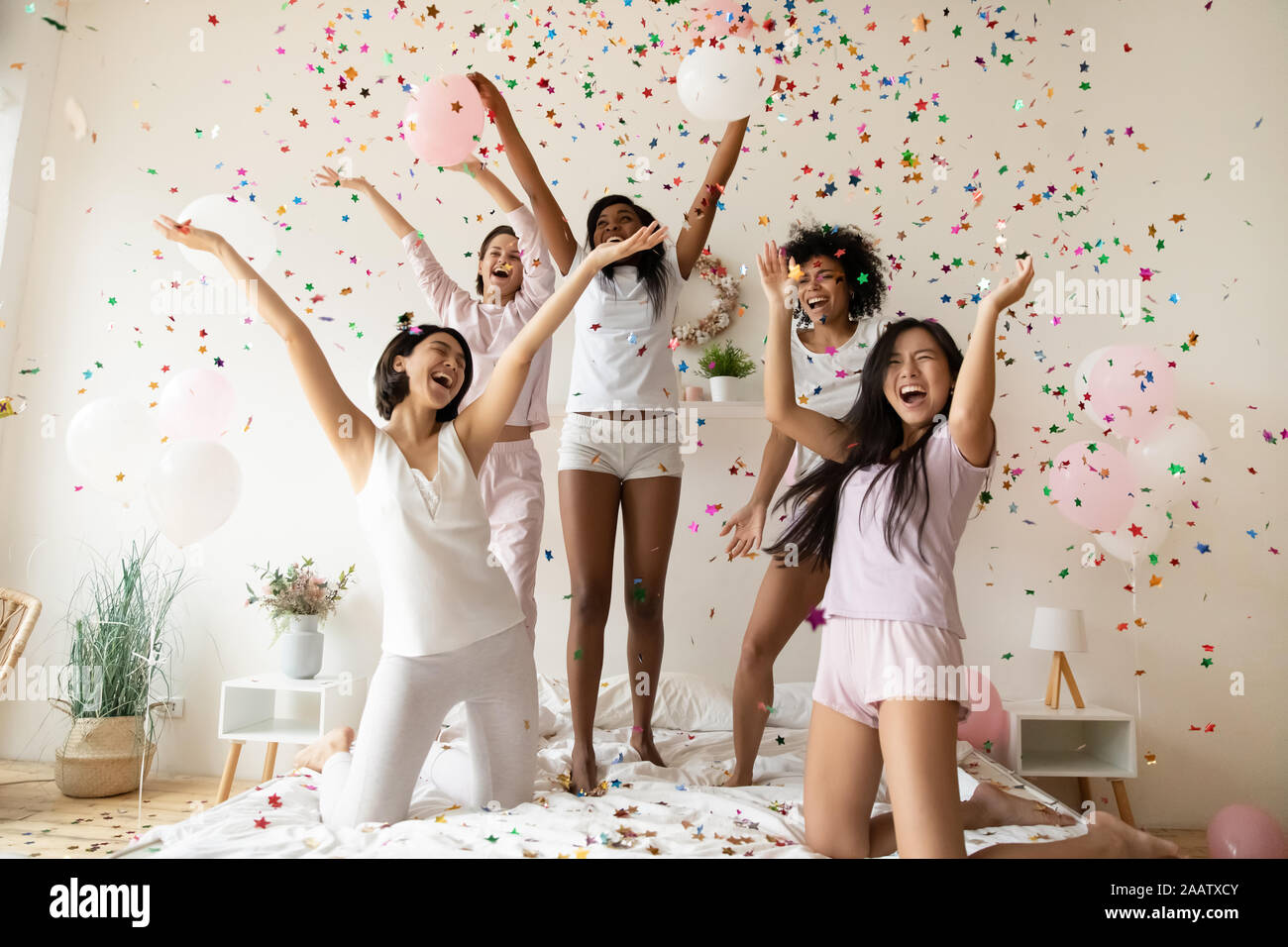 Excited multiethnic women wear pyjamas dance on bed in confetti Stock ...