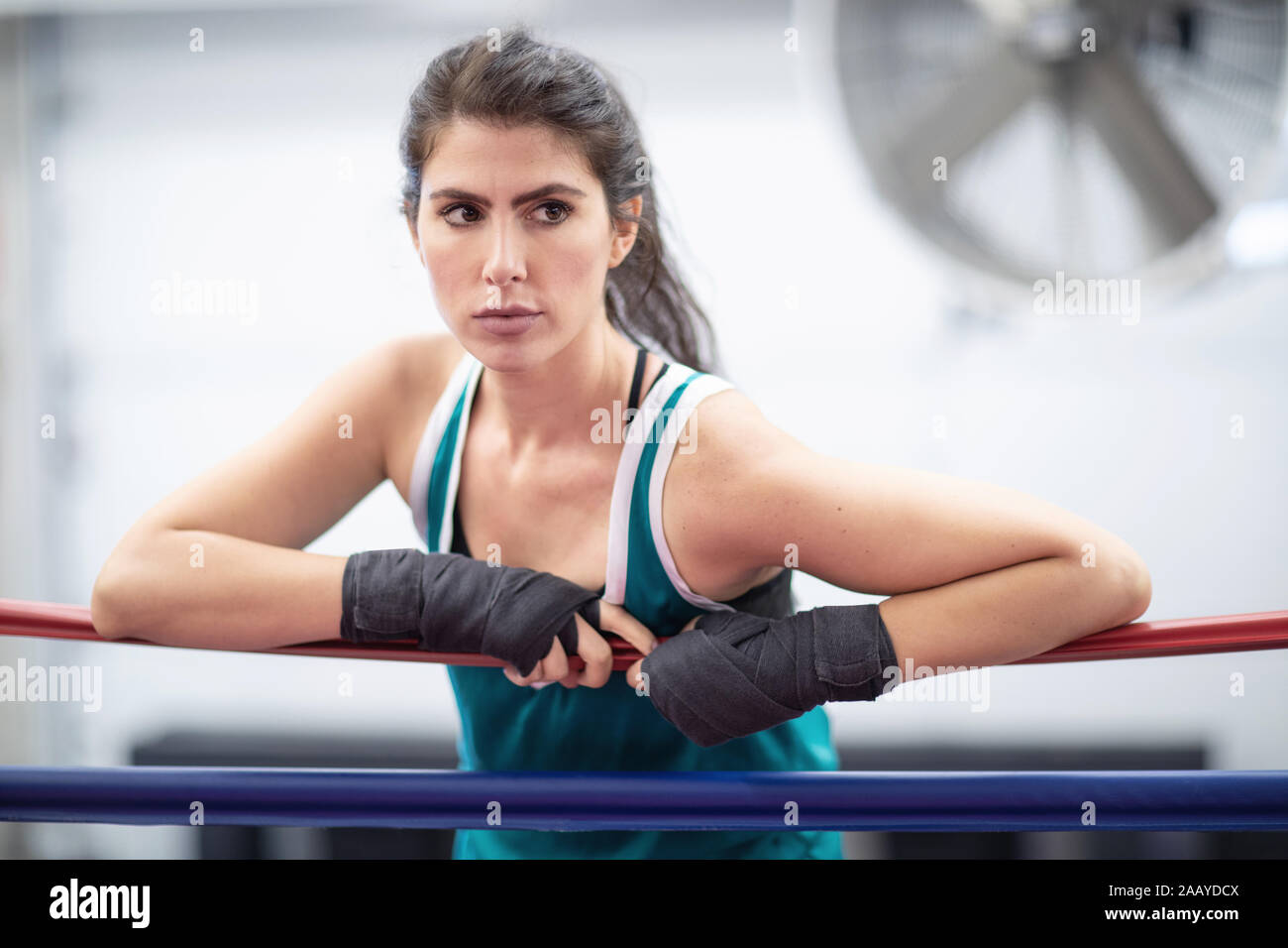 A badass female boxer is resting on ring ropes with her wraps on in a ...