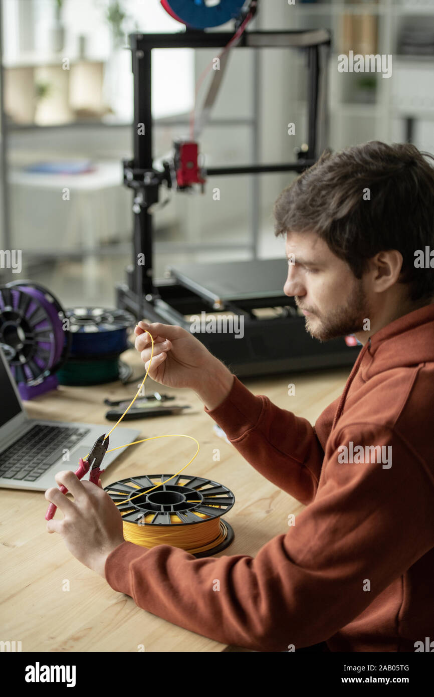 Creative man sitting by table and cutting piece of yellow filament ...