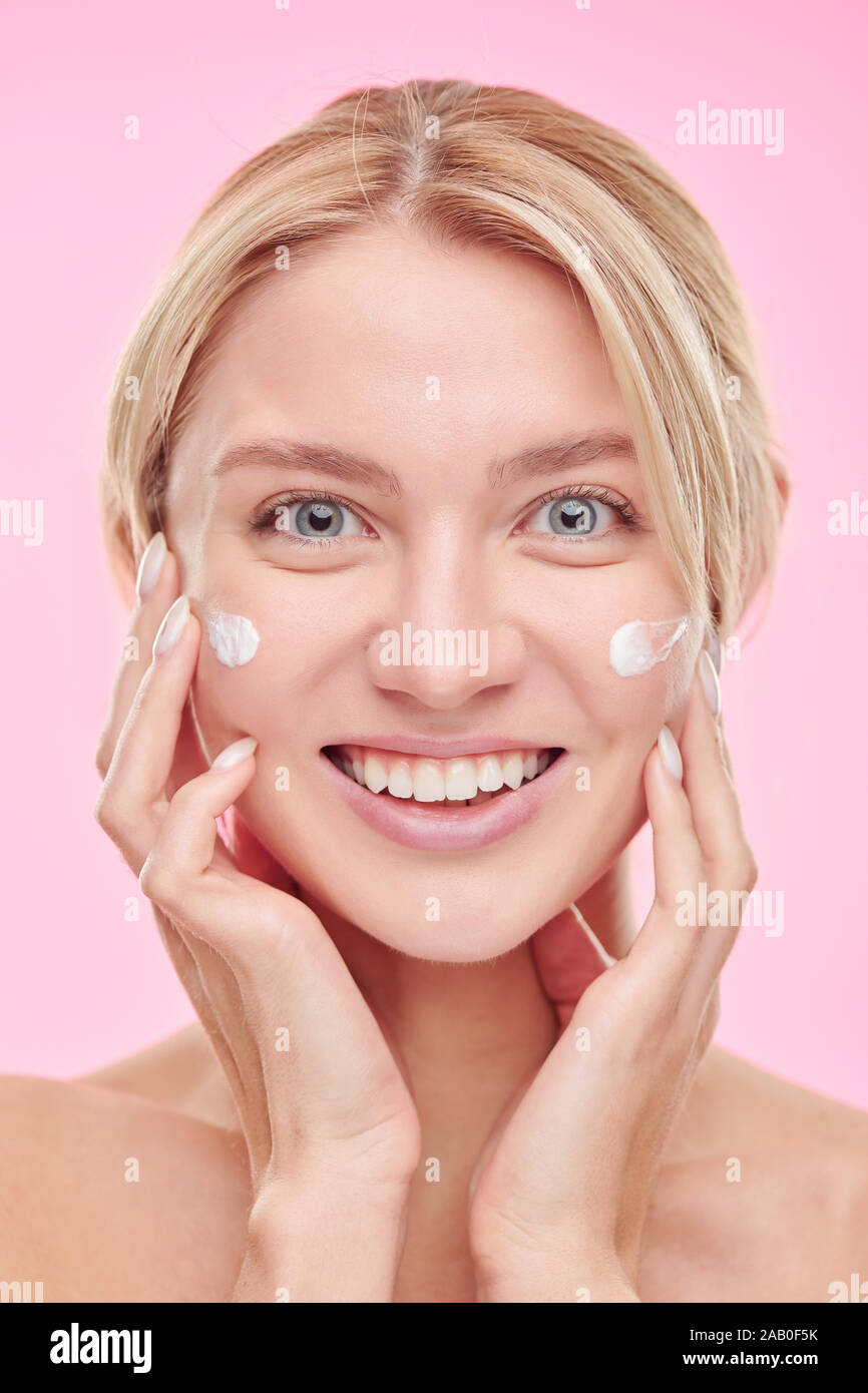 Young cheerful woman with toothy smile pampering cream on her face ...