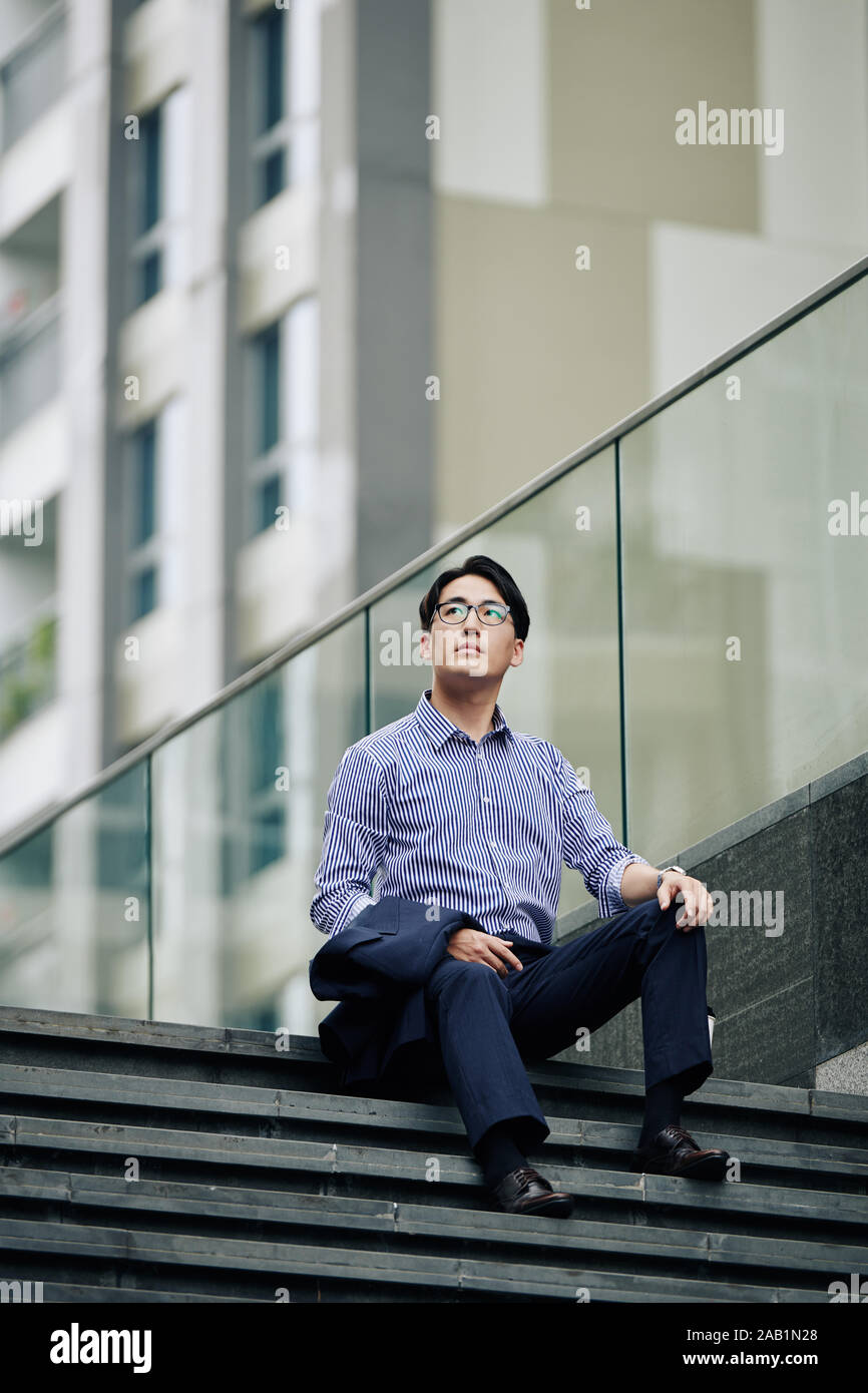 Dreamy Asian entrepreneur taking off jacket and resting on steps and ...