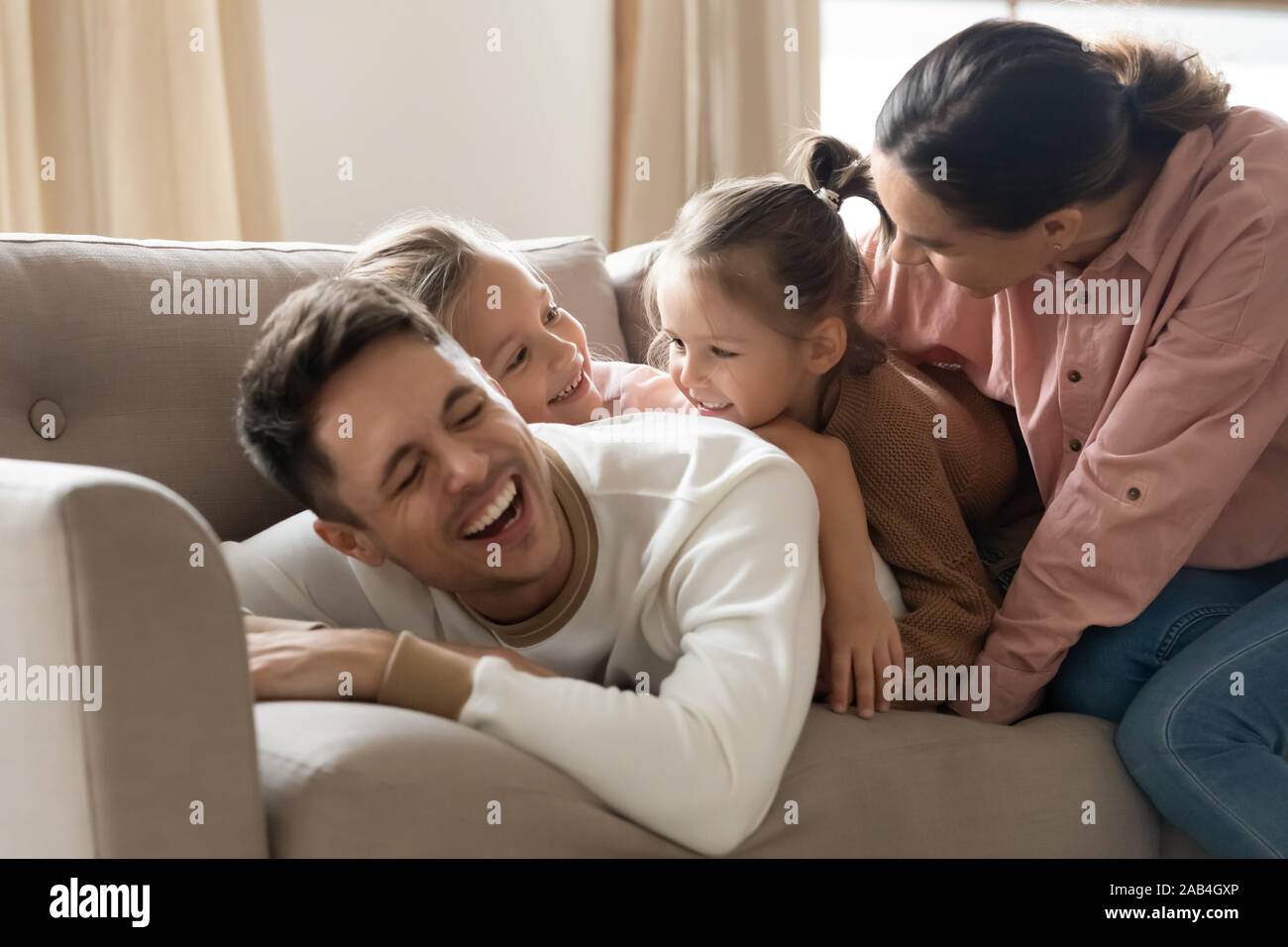 Cheerful parents laughing playing with small daughters lying on couch ...