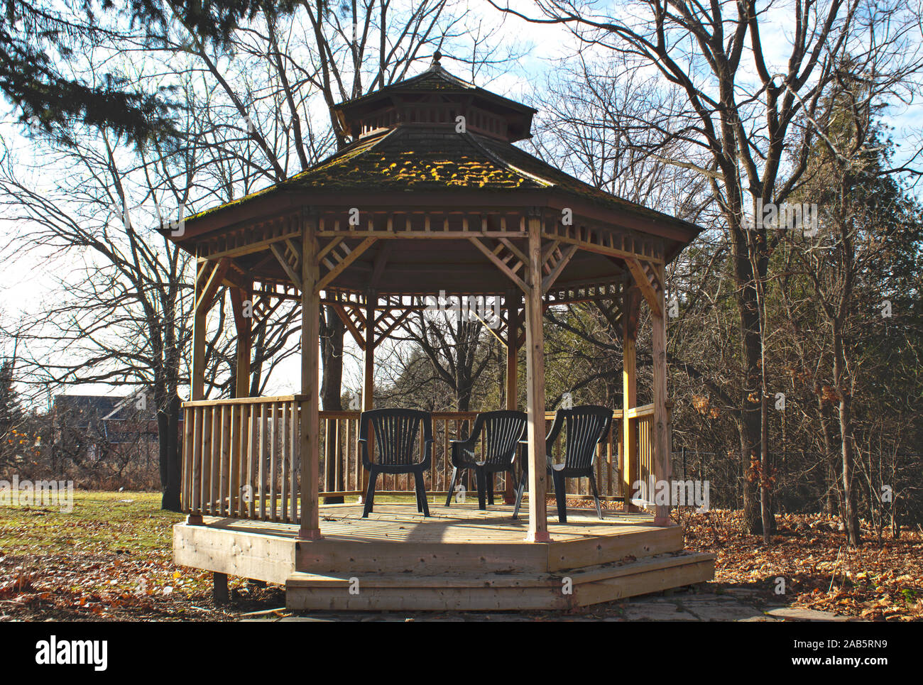 A wooden gazebo in fall, trees in the background, three seats in gazebo ...
