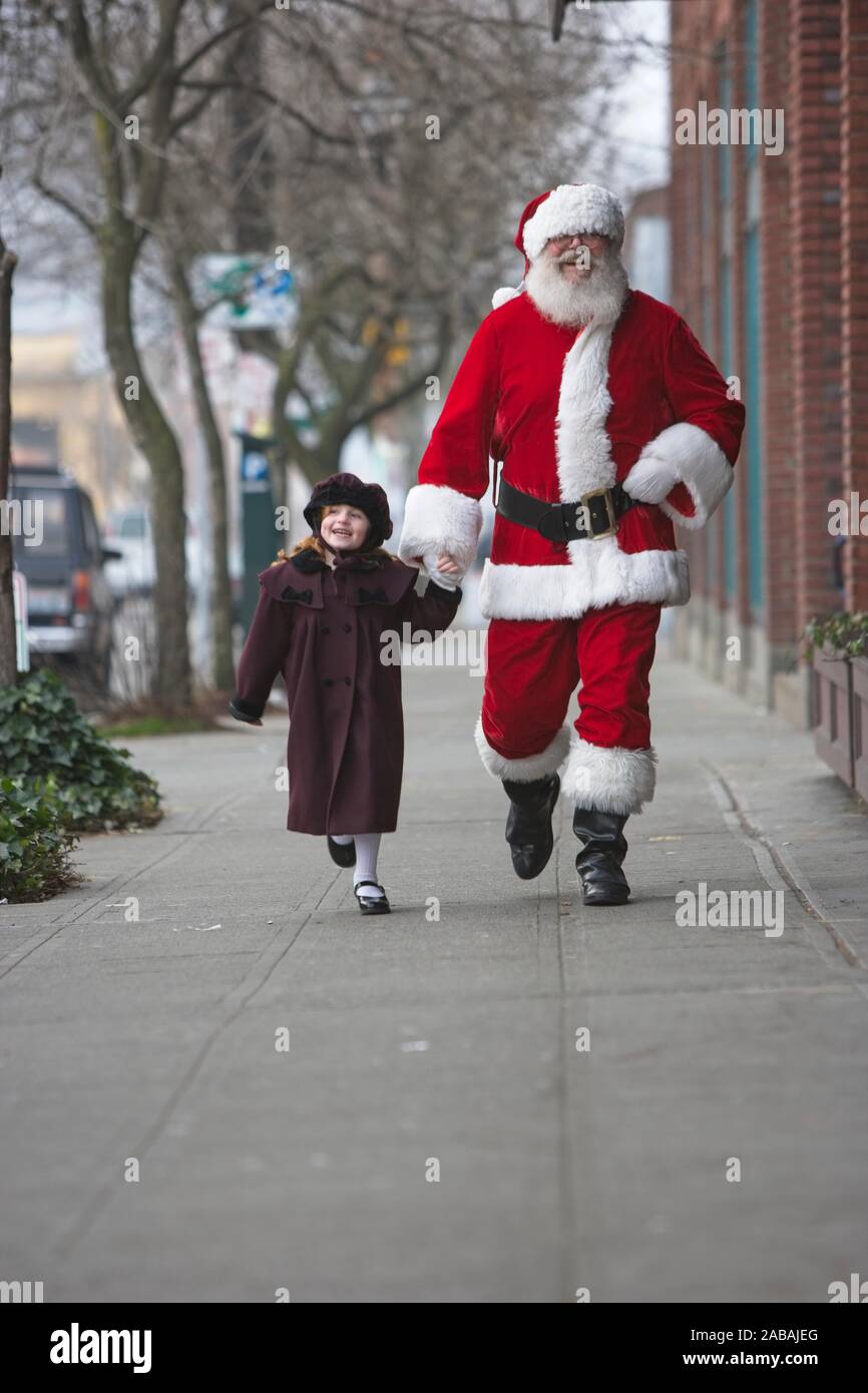 Santa Claus walking down a city sidewalk holding hands with a cute ...