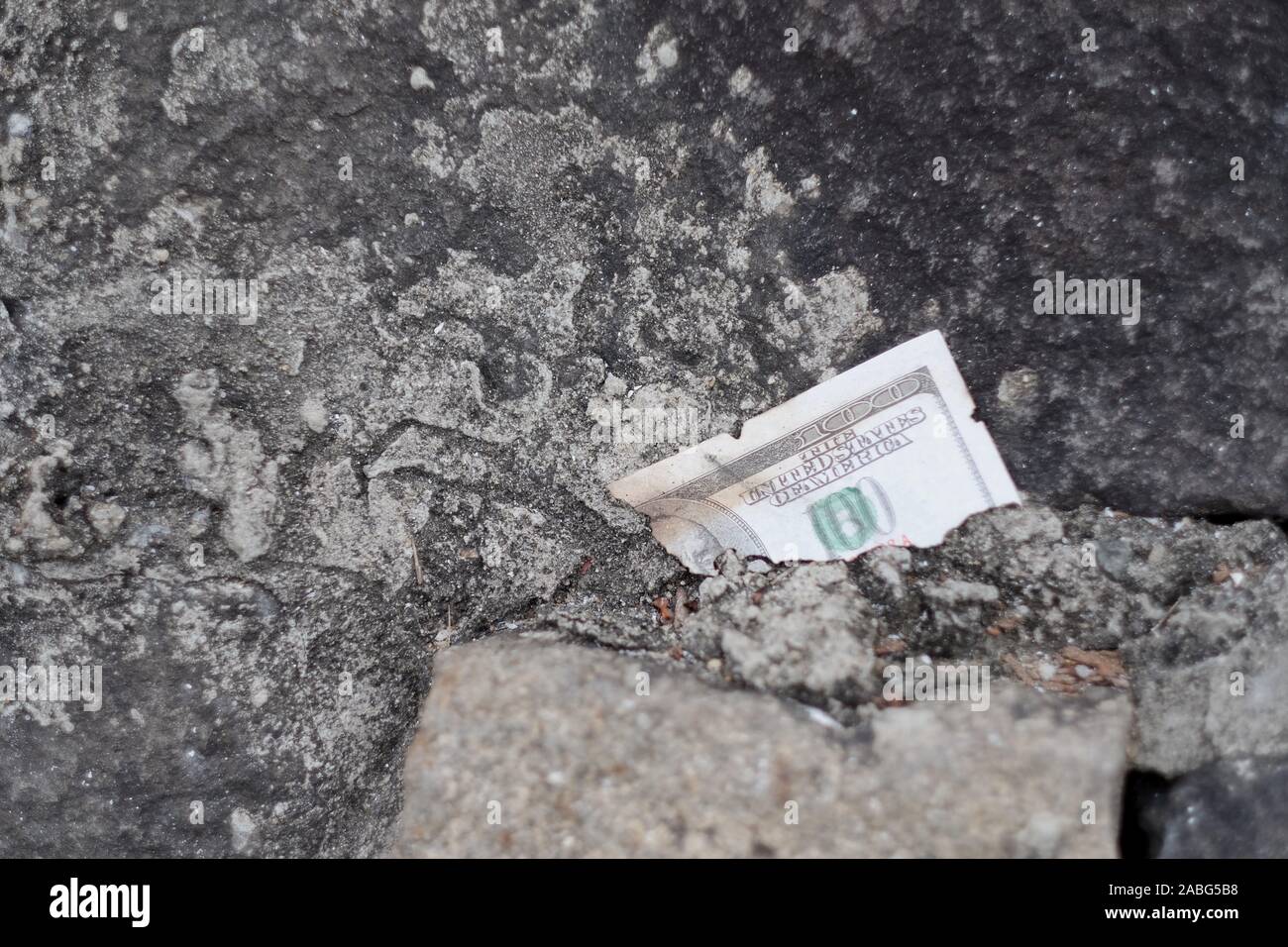 A piece of a hundred-dollar bill lies in a slot on an asphalt path ...