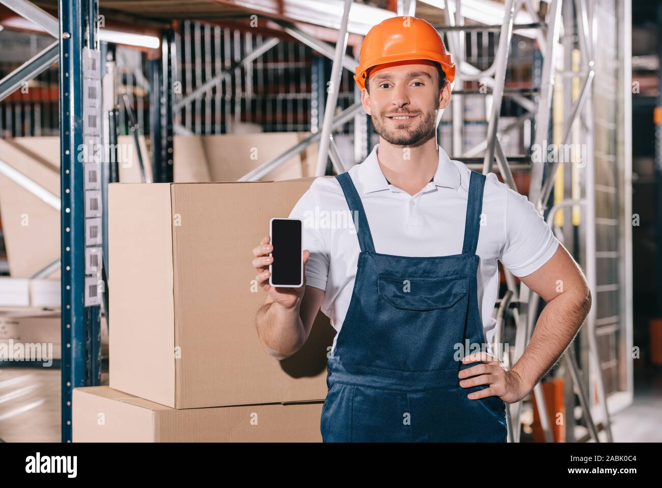 smiling loader standing with hand on hip and showing smartphone with ...