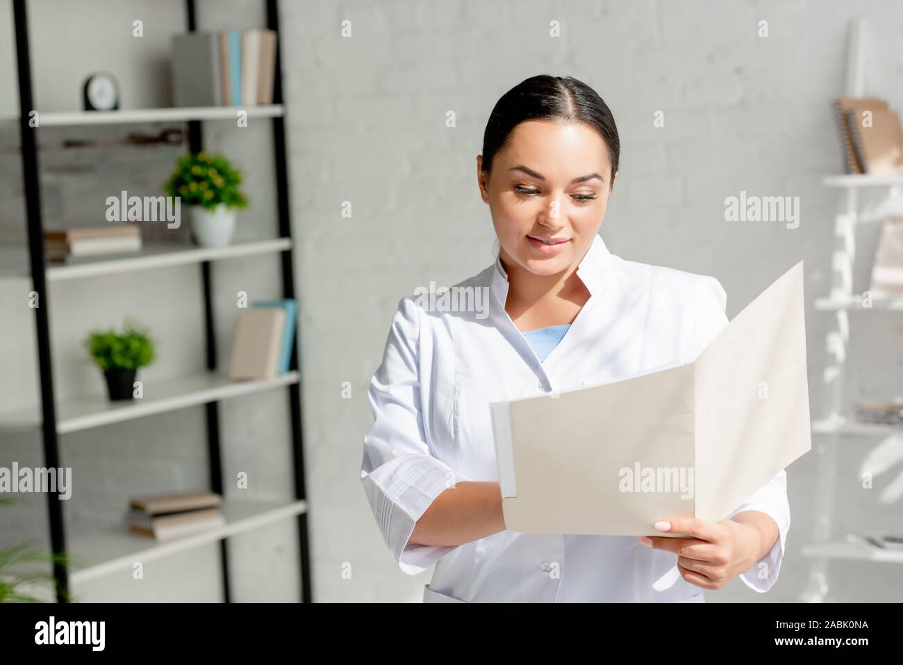 attractive and smiling dermatologist in white coat holding folder in ...