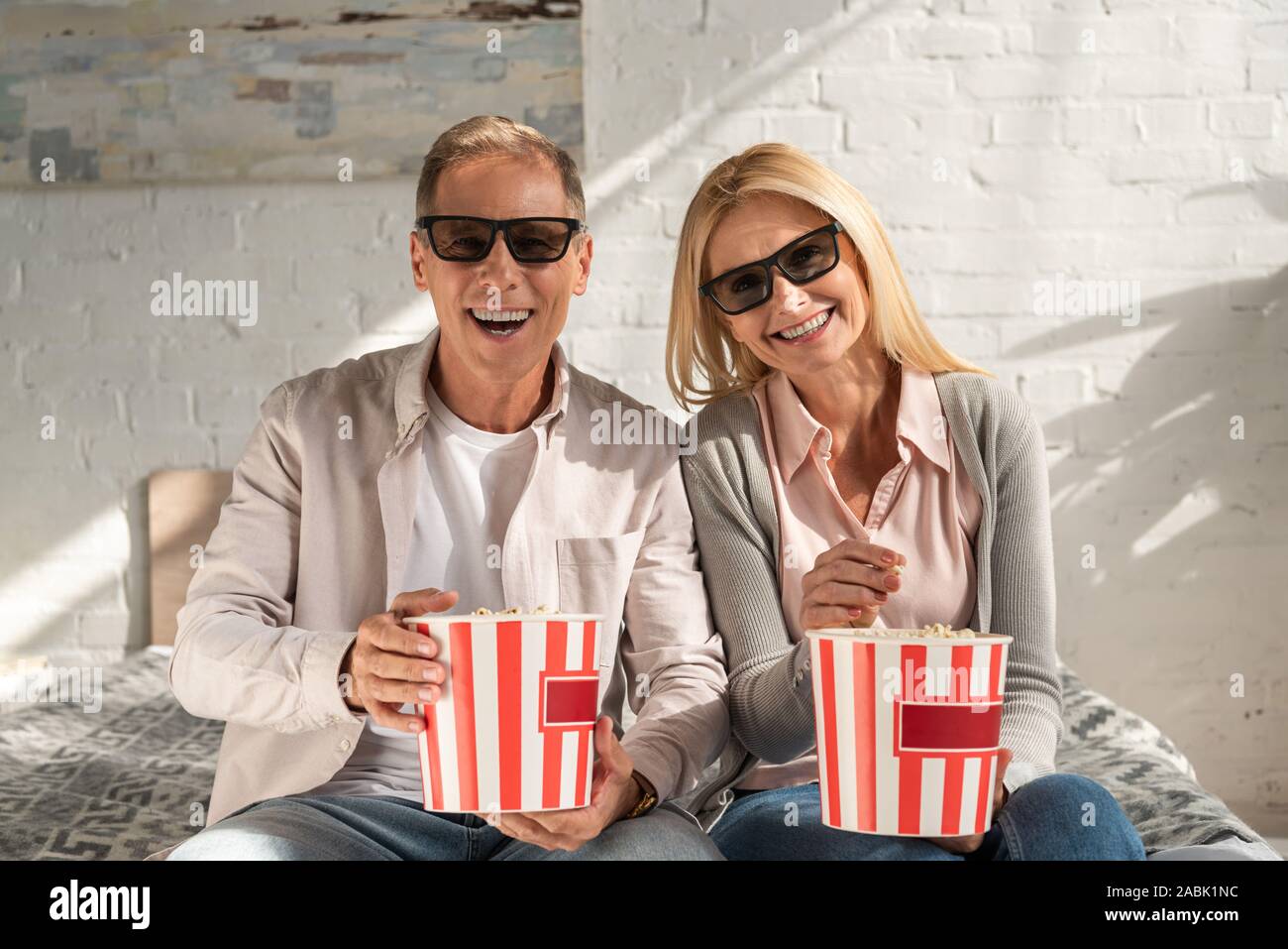 Smiling couple in 3d glasses holding buckets with popcorn on bed Stock Photo - Alamy