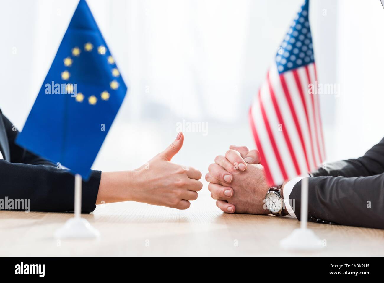 cropped view of diplomat of european union showing thumb up near ambassador of usa sitting with ...