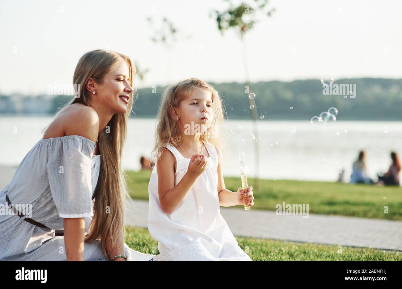 Photo of young mother and her daughter having good time on the green grass with lake at ...