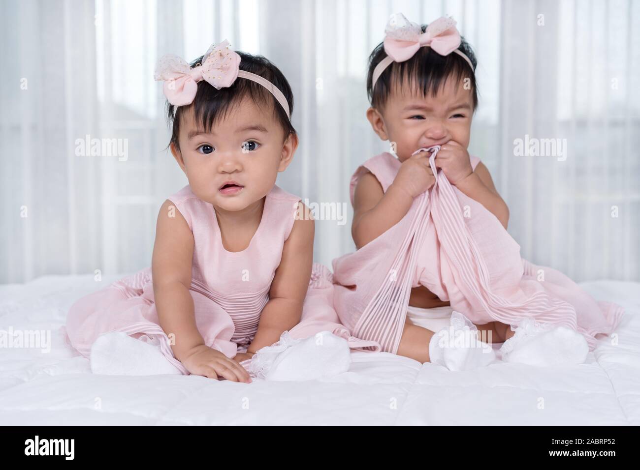 two twin babies in pink dress on a bed, one looking, one crying Stock ...