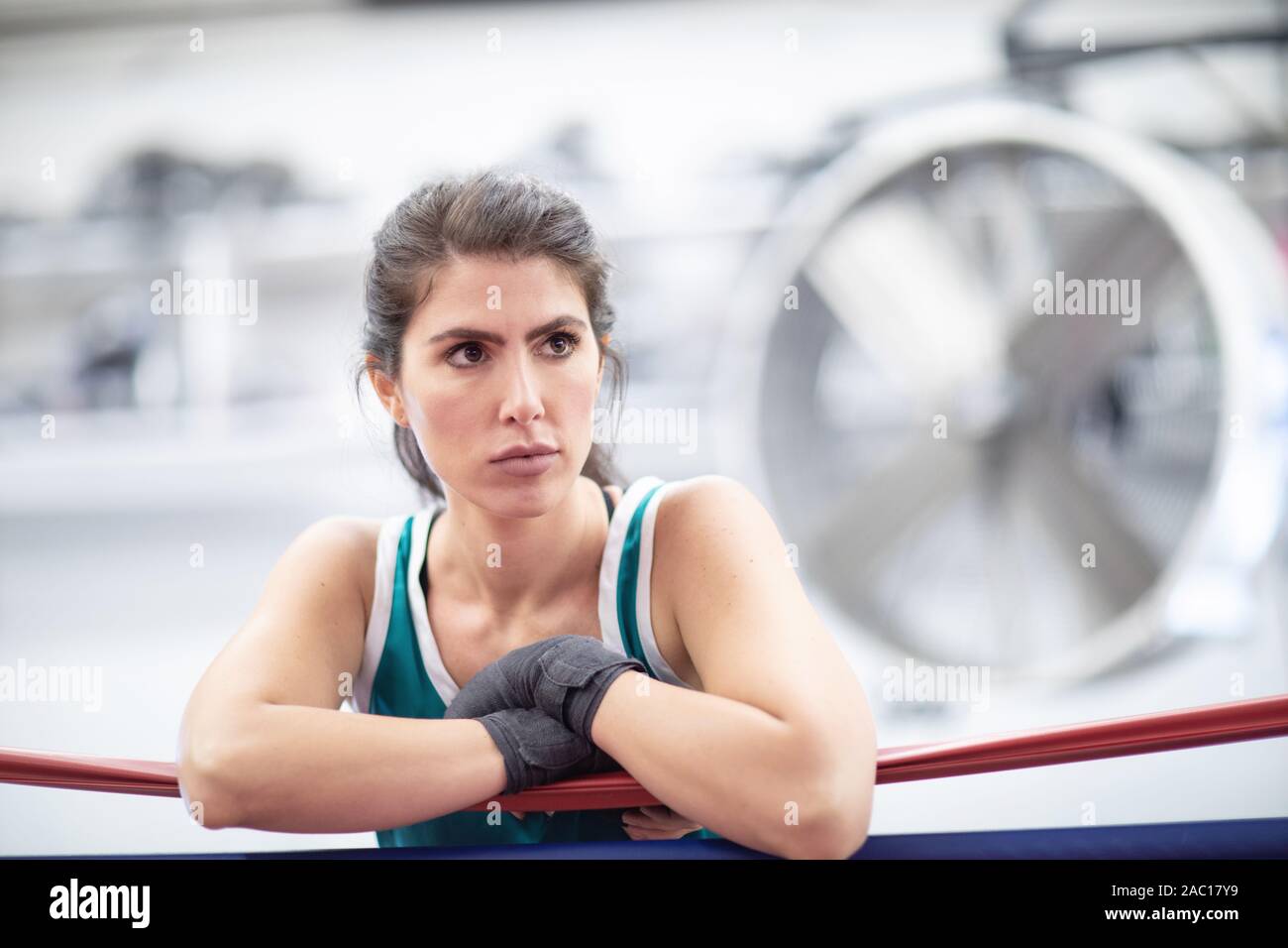 A badass female boxer is resting on ring ropes with her wraps on in a ...