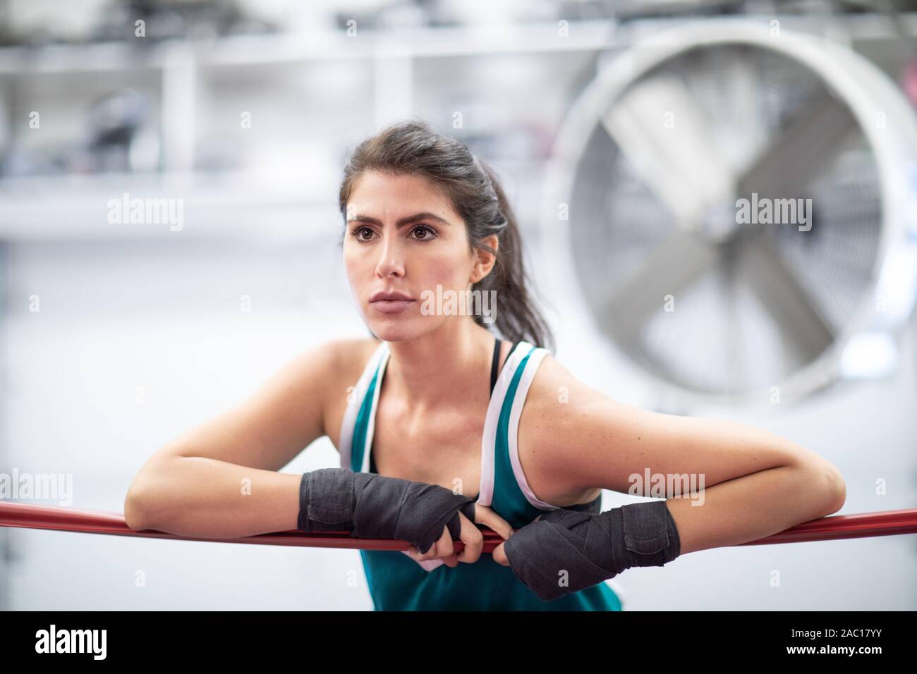 A badass female boxer is resting on ring ropes with her wraps on in a ...