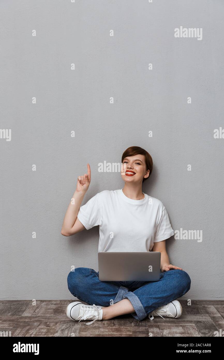 Image of young woman pointing finger upward while sitting on floor with ...