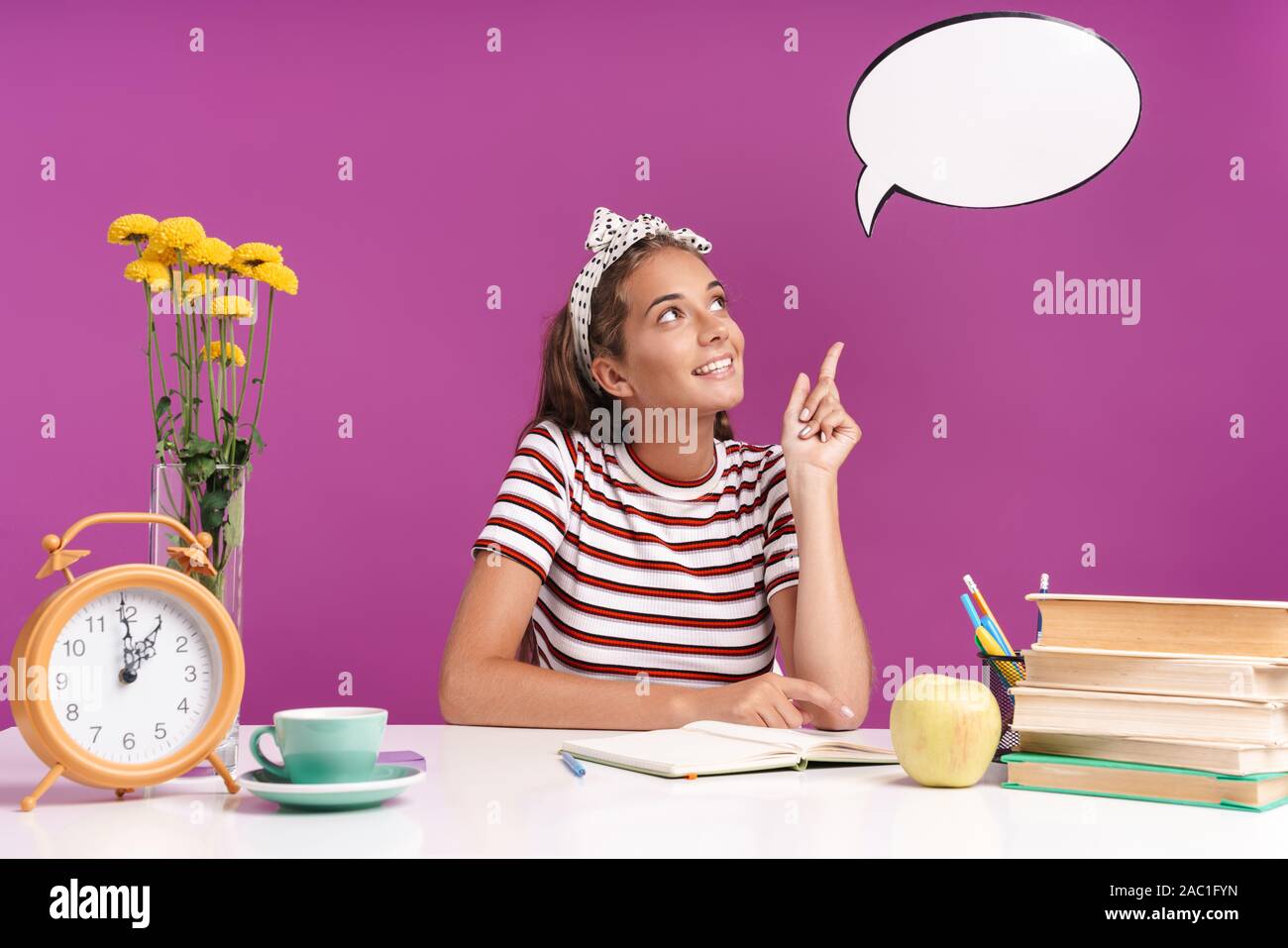 Attractive smiling young girl sitting at the desk isolated over pink ...