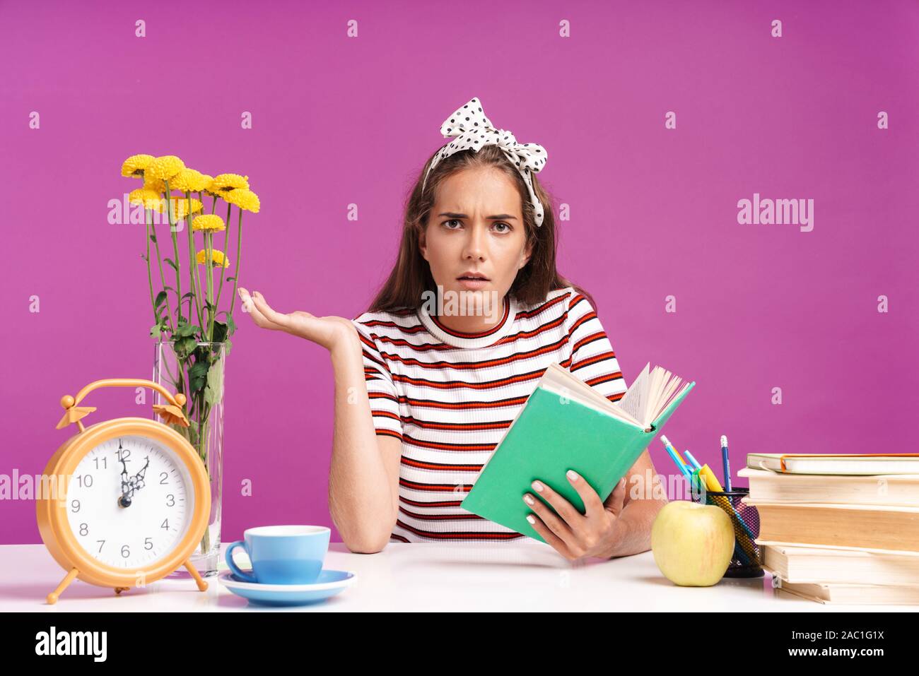 Attractive confused young girl sitting at the desk isolated over pink ...