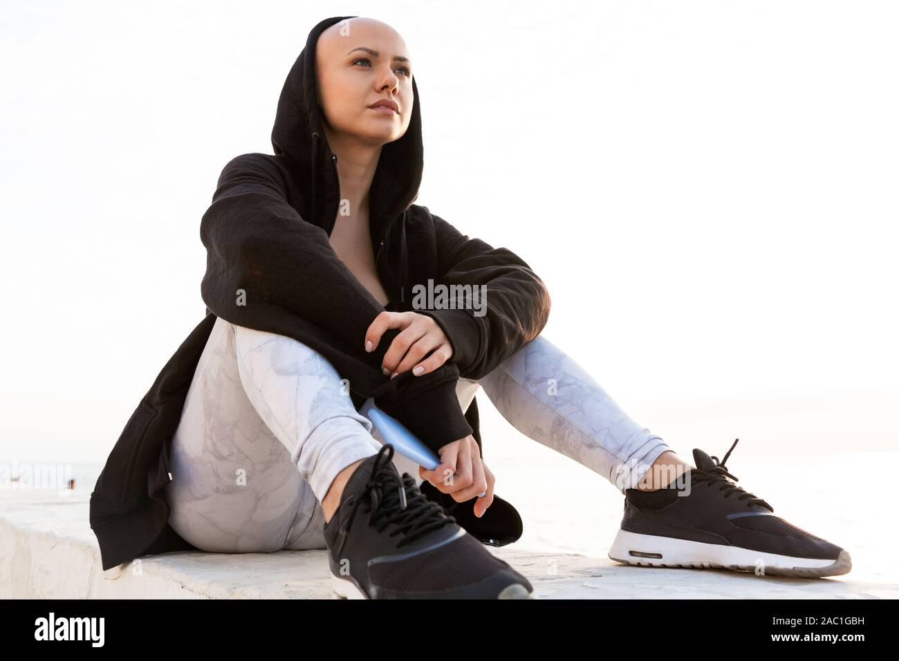 Image of focused bald woman in black hood using cellphone while sitting ...