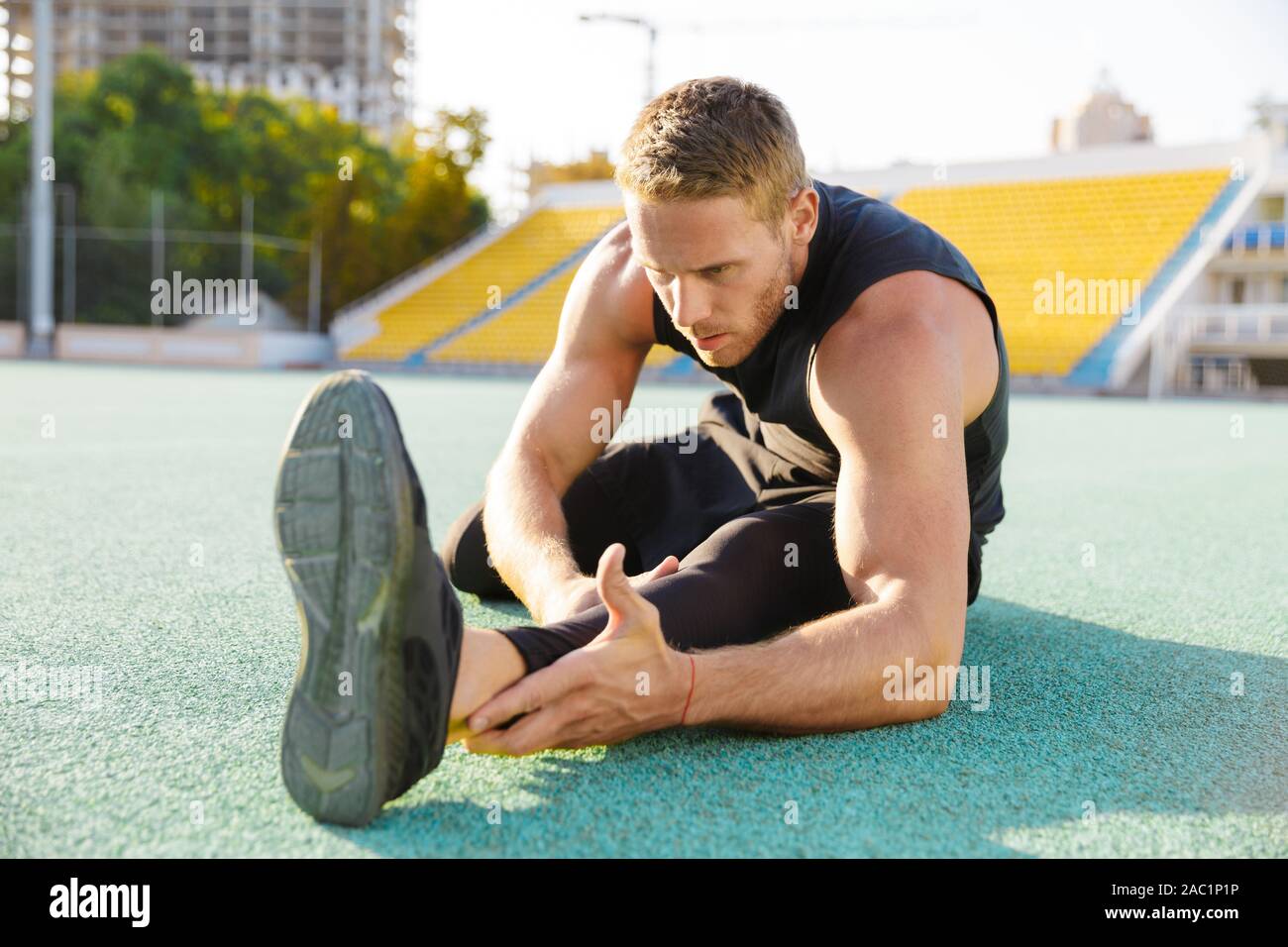 Image of concentrated man stretching his body at sports ground while ...