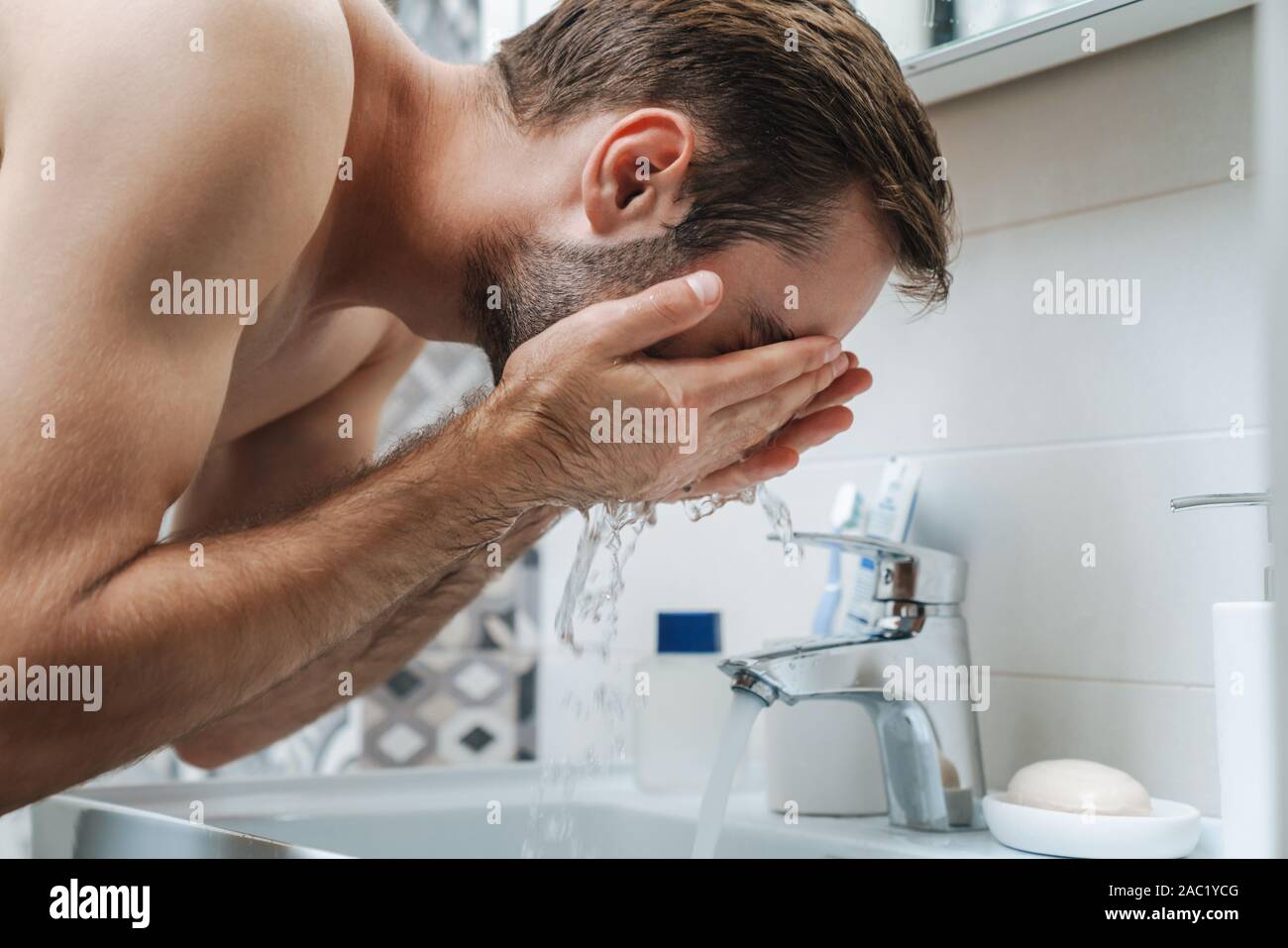 Handsome young shirtless man washing his face while standing at the ...