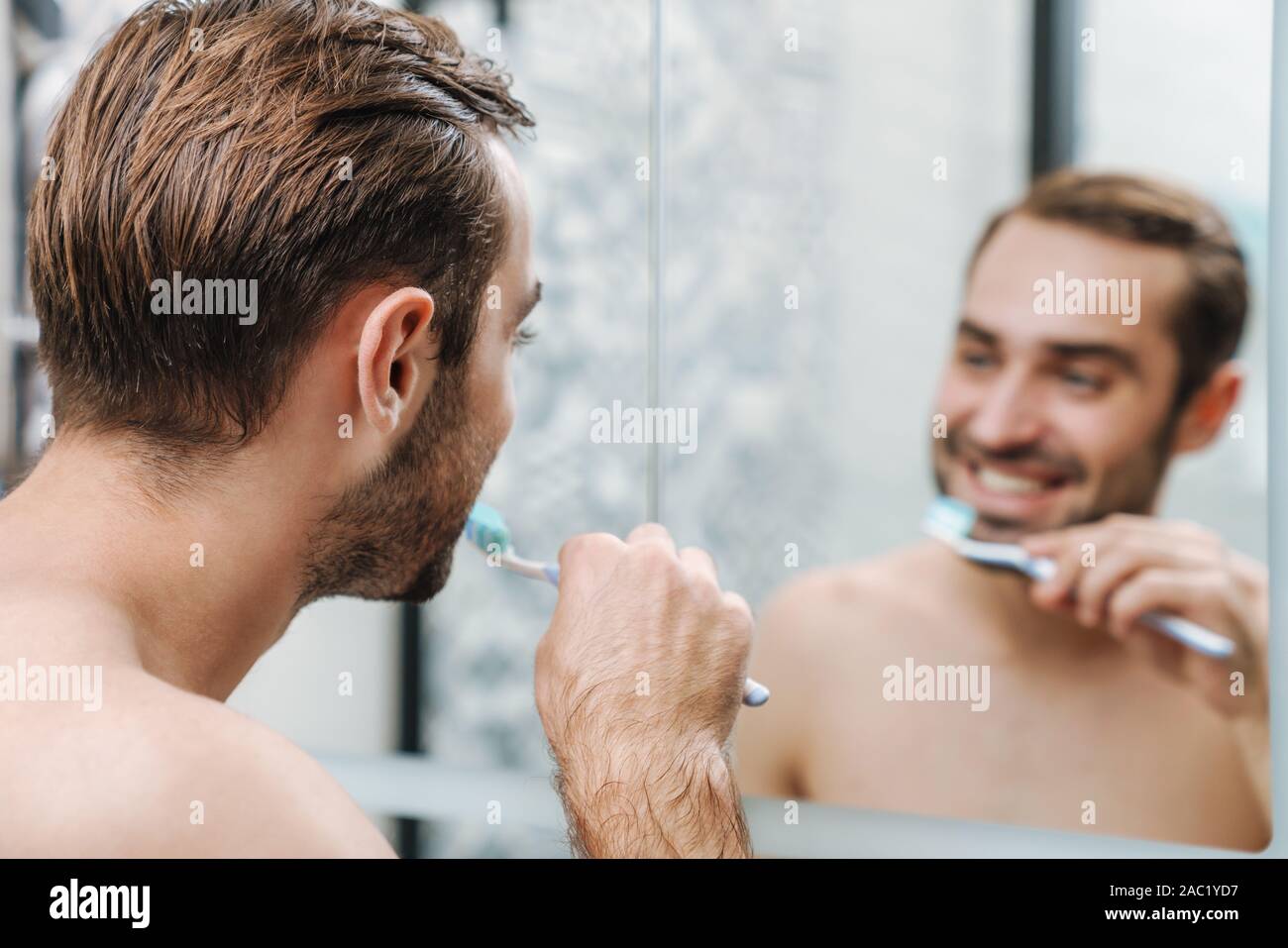 Attractive smiling shirtless man brushing teeth while standing at the ...