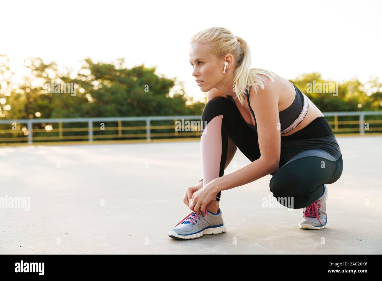 Image of blonde woman wearing tracksuit and earpods tying her sneakers ...