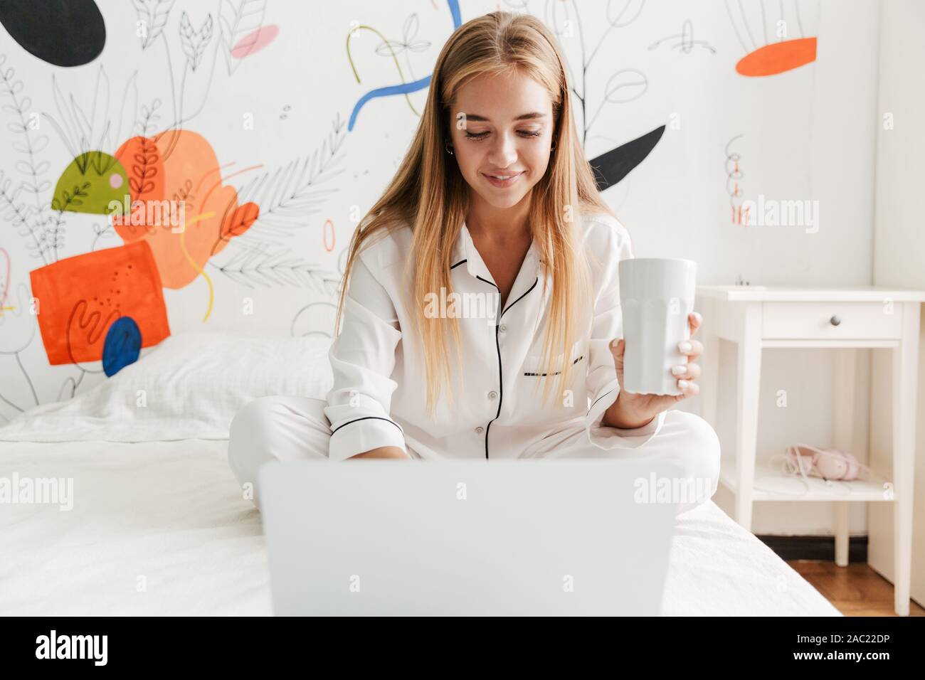 Image of a cheerful young cute girl in pajama at home on bed using ...