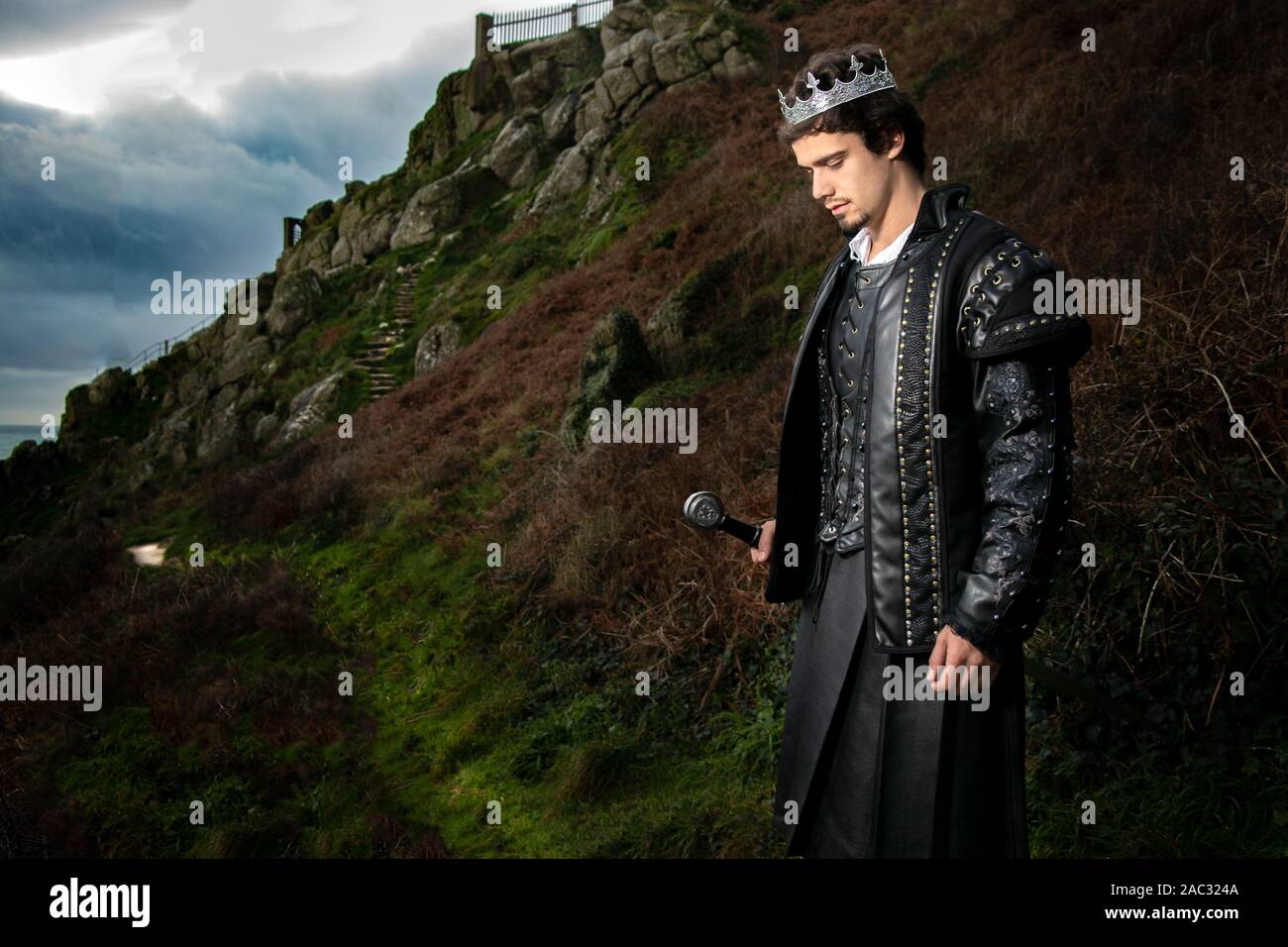 Handsome king with sword stands in contemplation with hill and parts of ...
