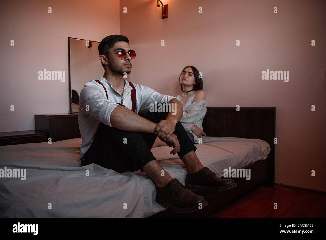 a young stylish guy in glasses sits on the edge of the bed with a ...
