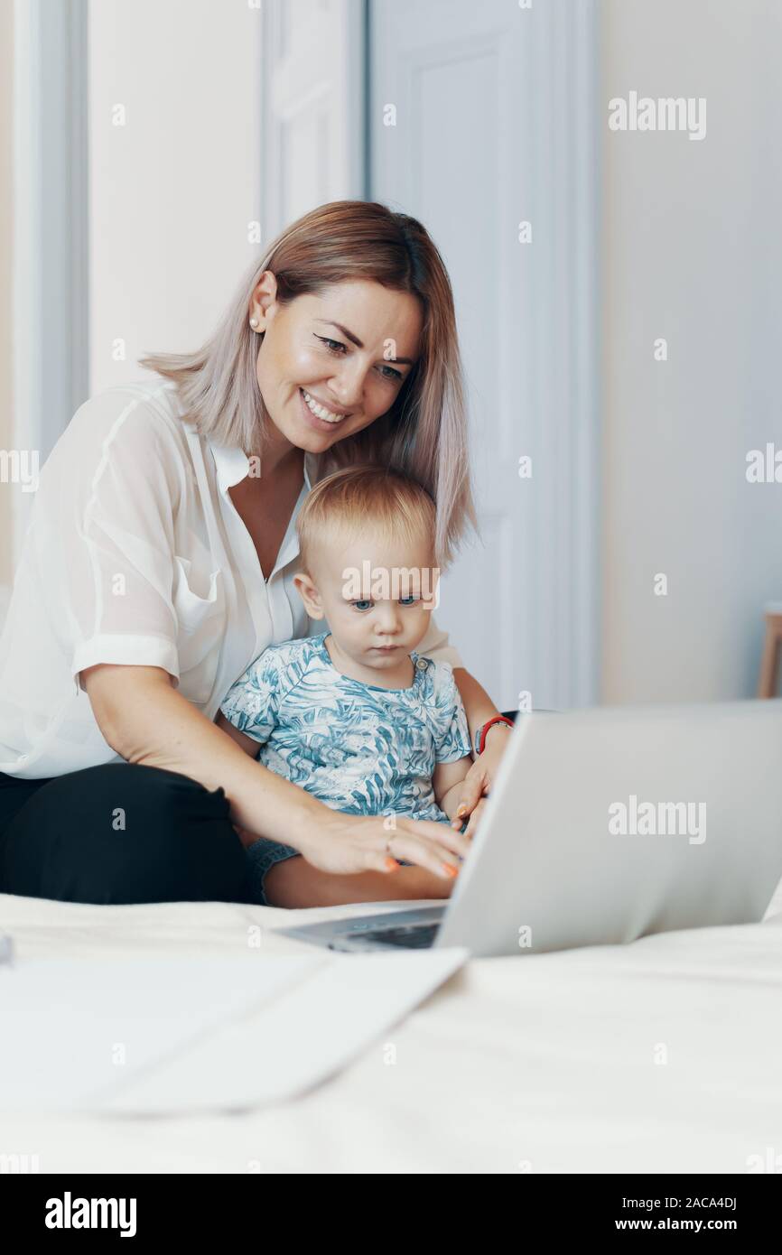 Young mother with her child working on laptop in bedroom at home. Multi-tasking, freelance and ...