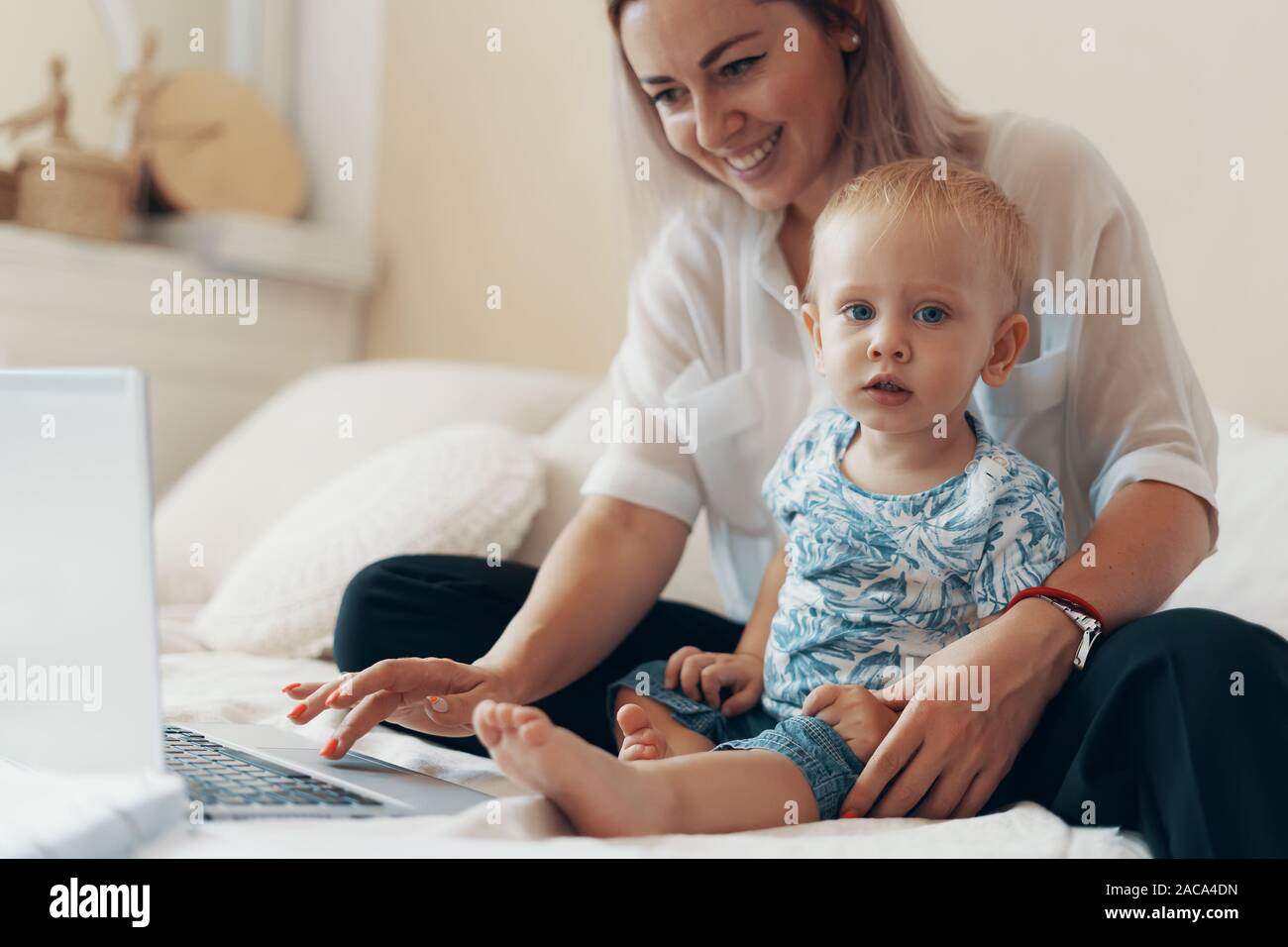 Young mother with her child working on laptop in bedroom at home. Multi-tasking, freelance and ...