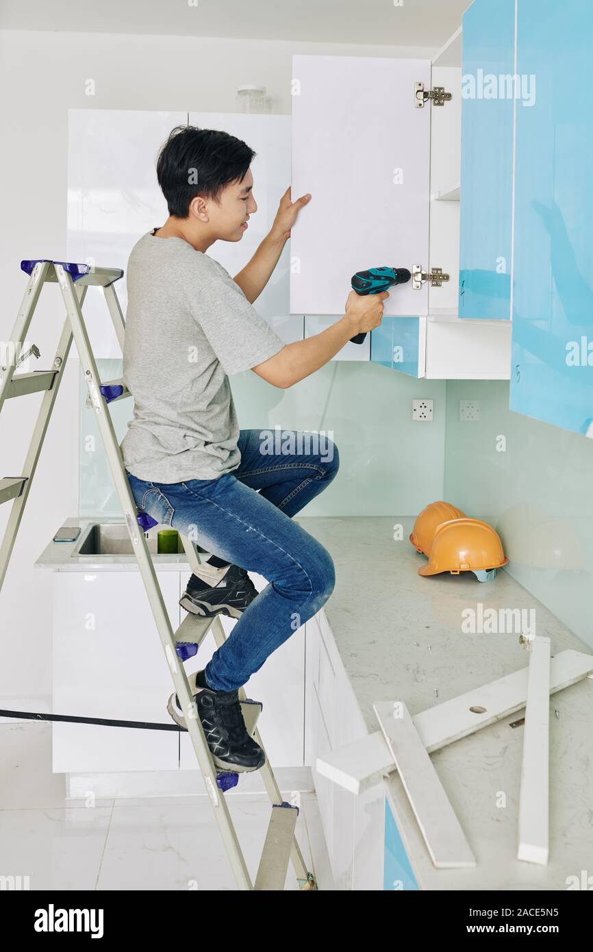 Young Asian man sitting on ladder when fixing door of kitchen cupboard ...