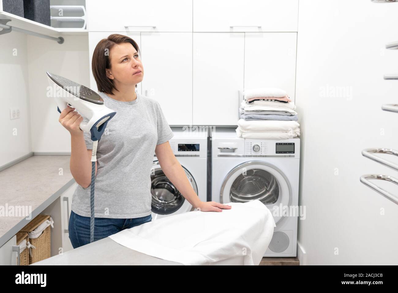 Woman ironing white shirt on board in laundry room with washing machine ...