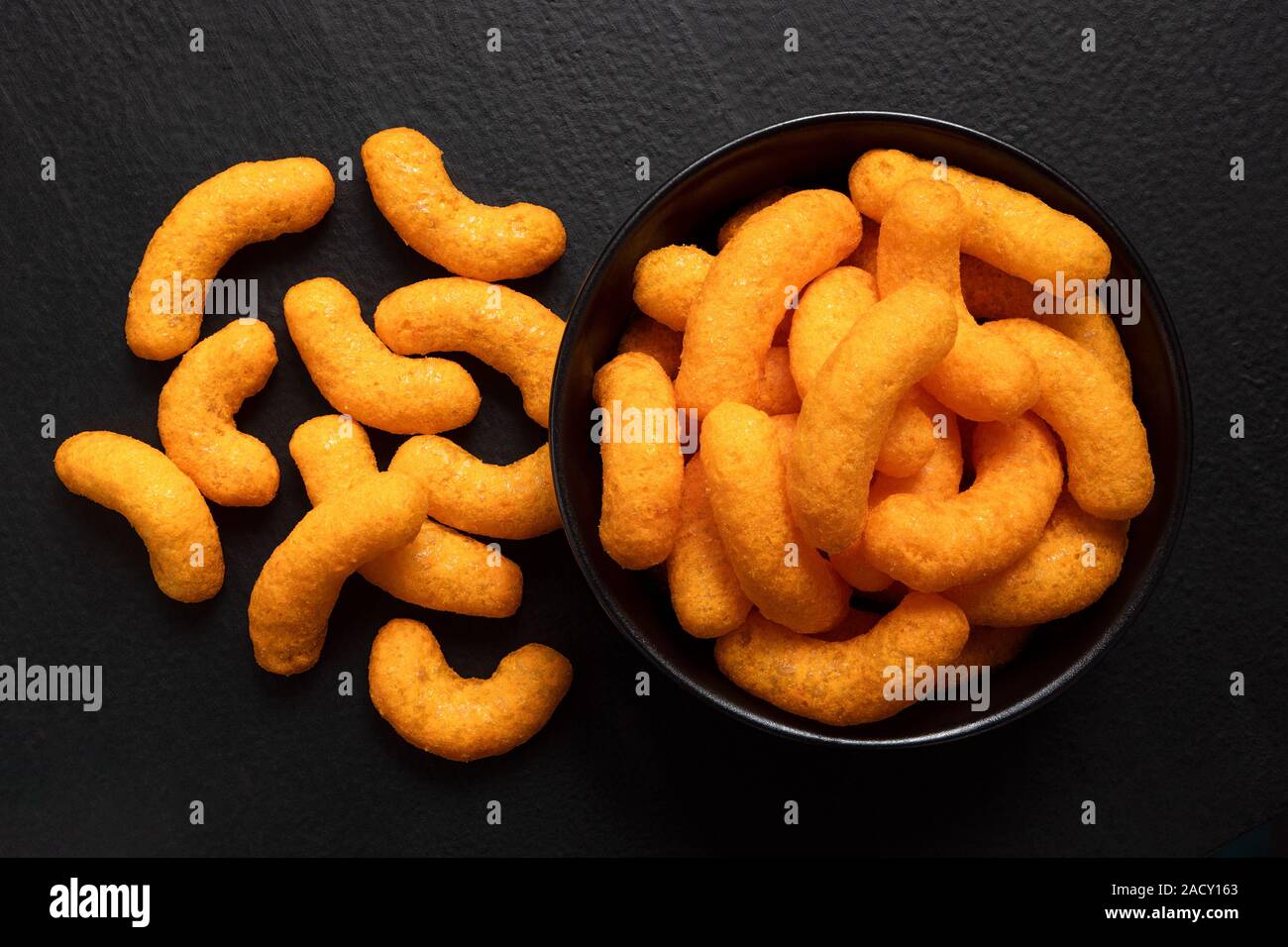 Extruded cheese puffs in a black ceramic bowl next to spilled cheese ...