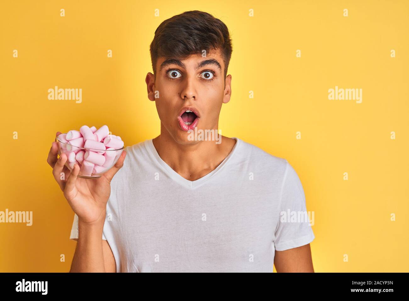 Young indian man holding bowl with marshmallows over isolated yellow ...