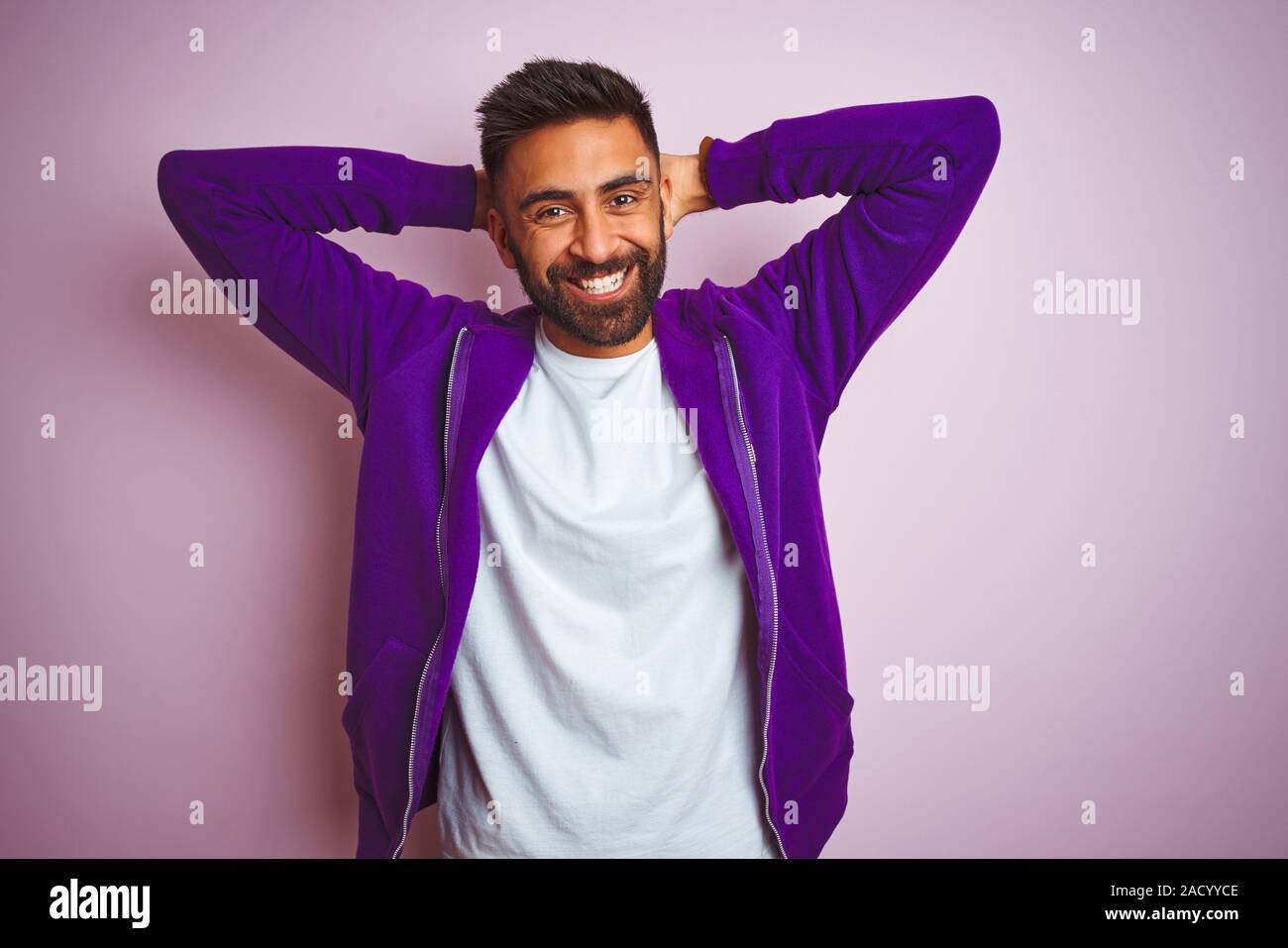 Young indian man wearing purple sweatshirt standing over isolated pink ...