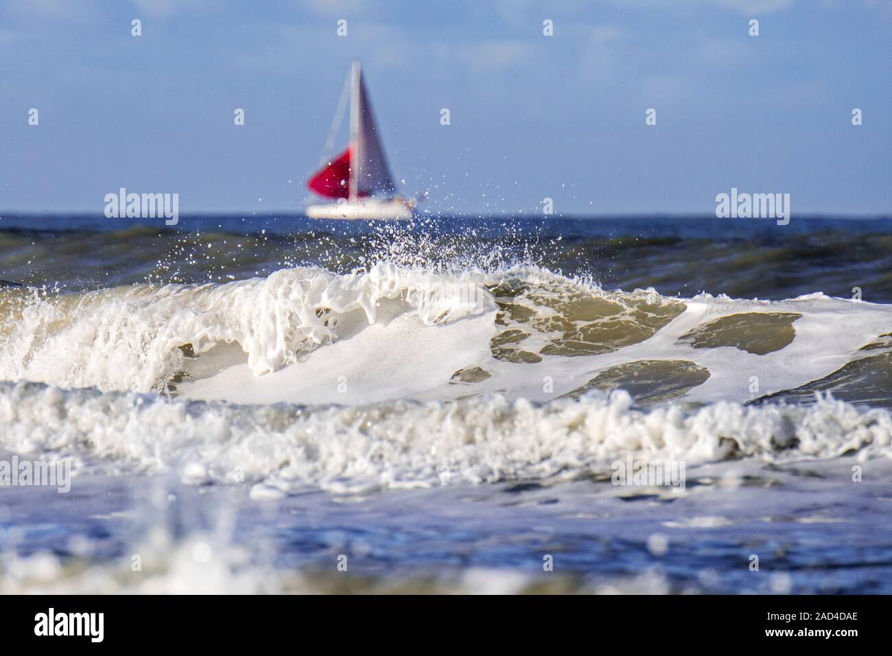 Wave breaking on the beach and sailing boat / sailboat with red sail at ...