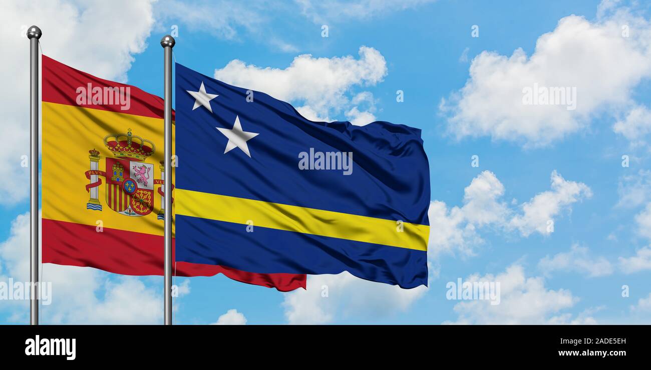 Spain and Curacao flag waving in the wind against white cloudy blue sky ...