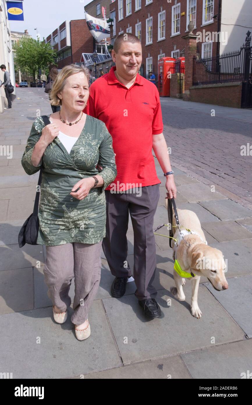 Vision impaired man with guide dog and sighted guide walking along a ...