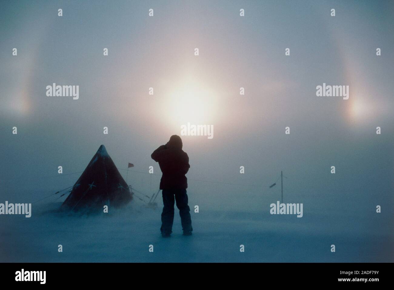 Sun-dogs. Silhouetted figure beside a tent watching sun-dogs over ...