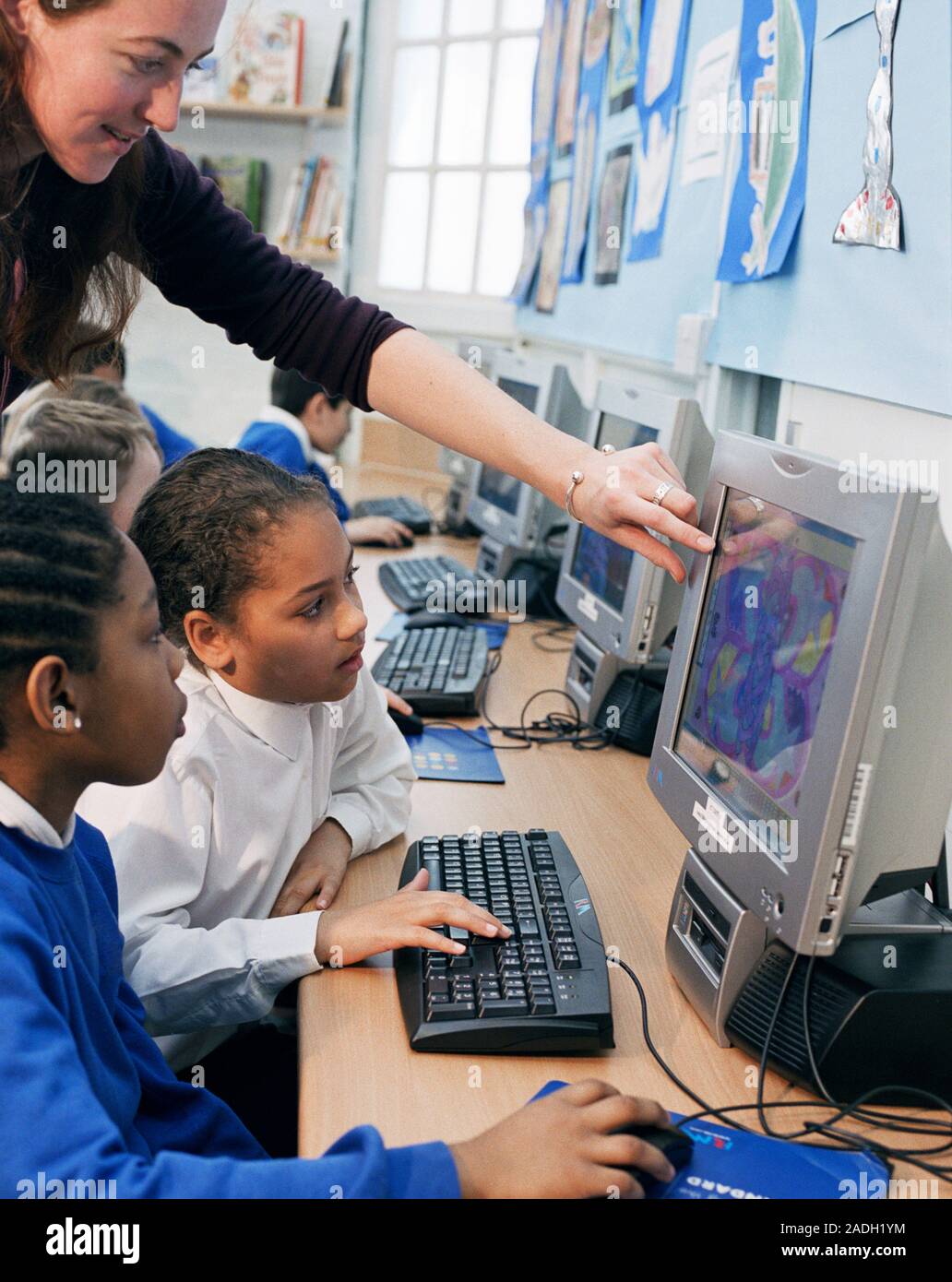 Schoolchildren using computers at school. The class teacher is pointing ...